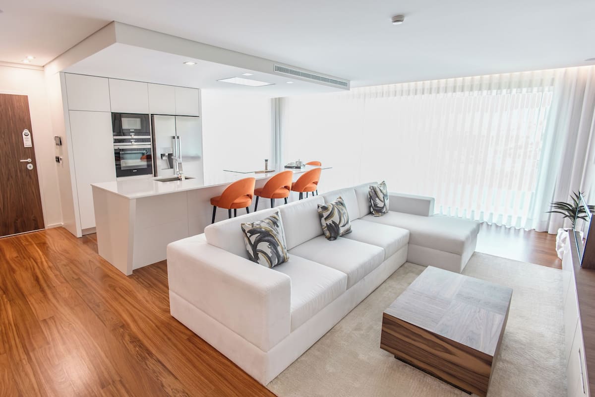 Uma sala de estar moderna com um sofá branco, uma mesa de centro de madeira e uma área de cozinha com bancada branca e cadeiras laranja.