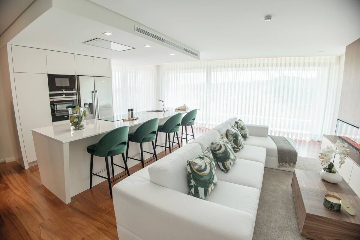 Cozinha moderna com ilha central, banquetas, sofá com almofadas decorativas e área de jantar com uma grande janela.