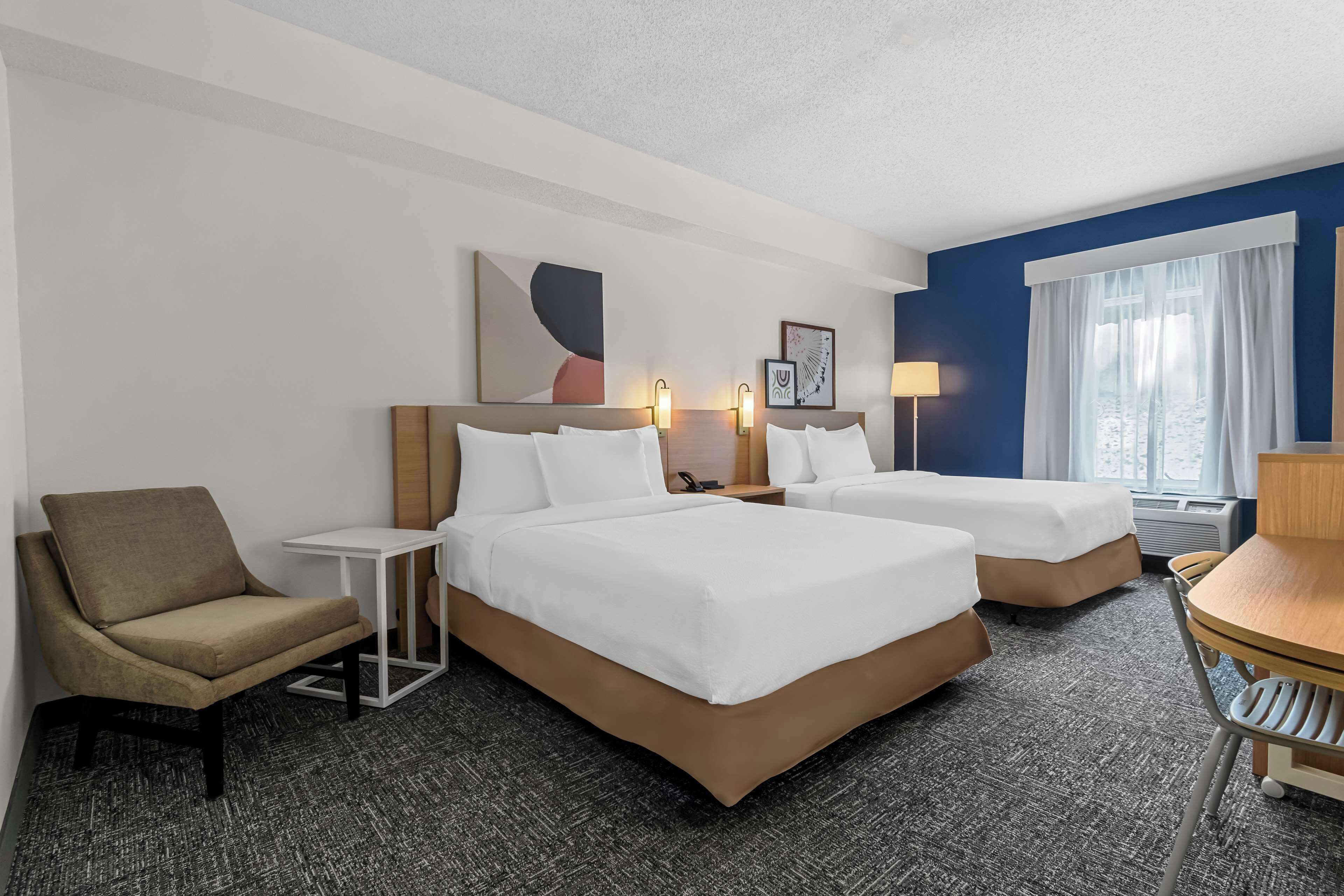Quarto de hotel com duas camas, uma mesinha, uma cadeira e uma escrivaninha.