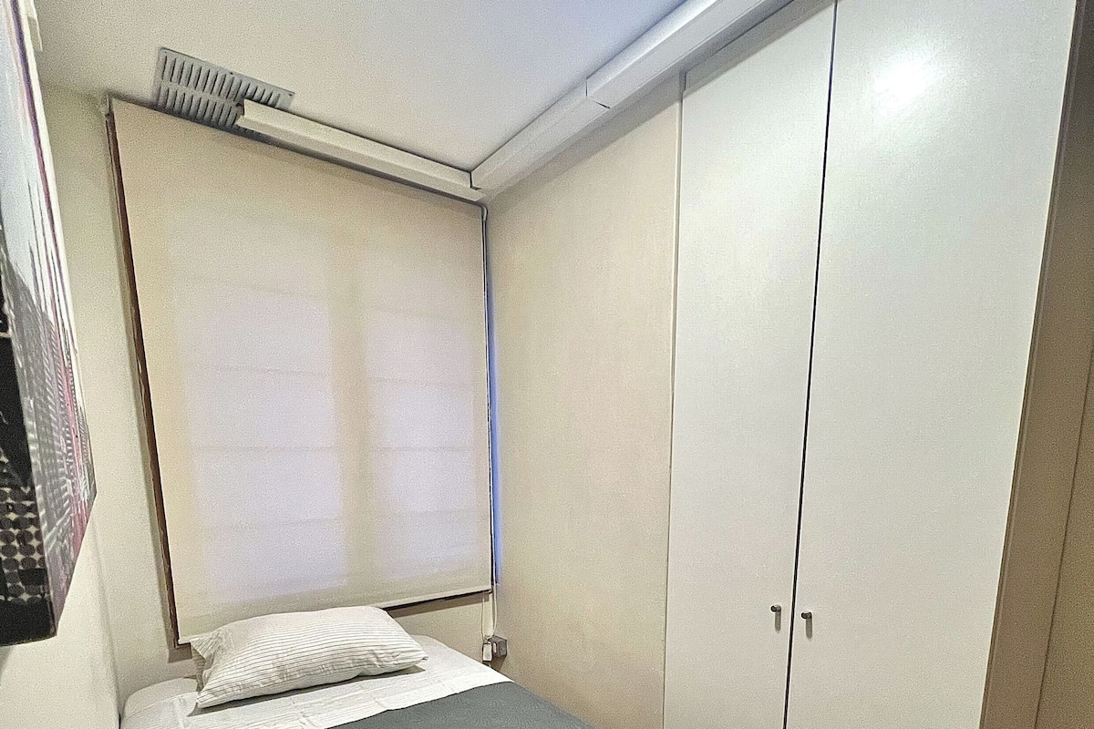 Quarto pequeno com uma cama de solteiro, um edredom cinza, um travesseiro branco, uma abertura no teto e um guarda-roupa com duas portas.