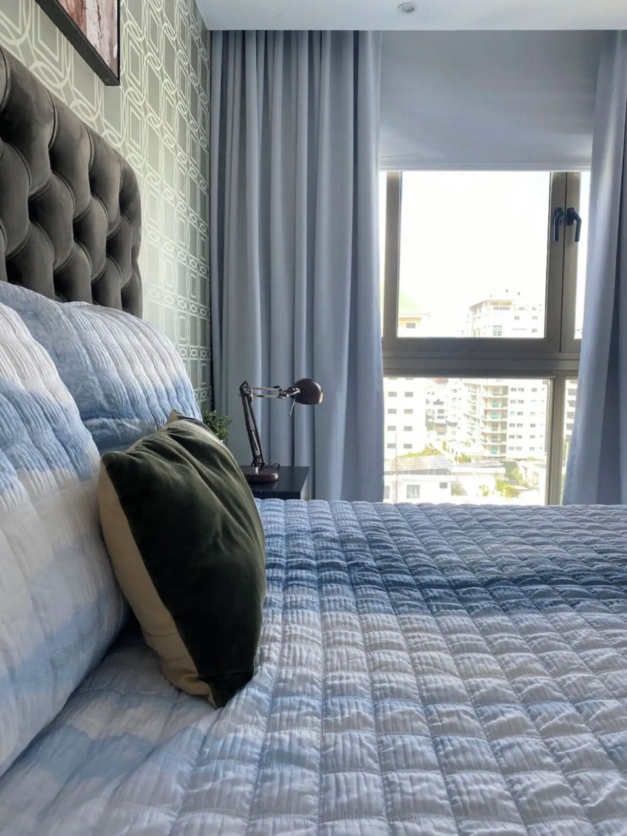 Quarto com cabeceira estofada, cama com colcha azul e branca, almofada verde e mesa de cabeceira com abajur.