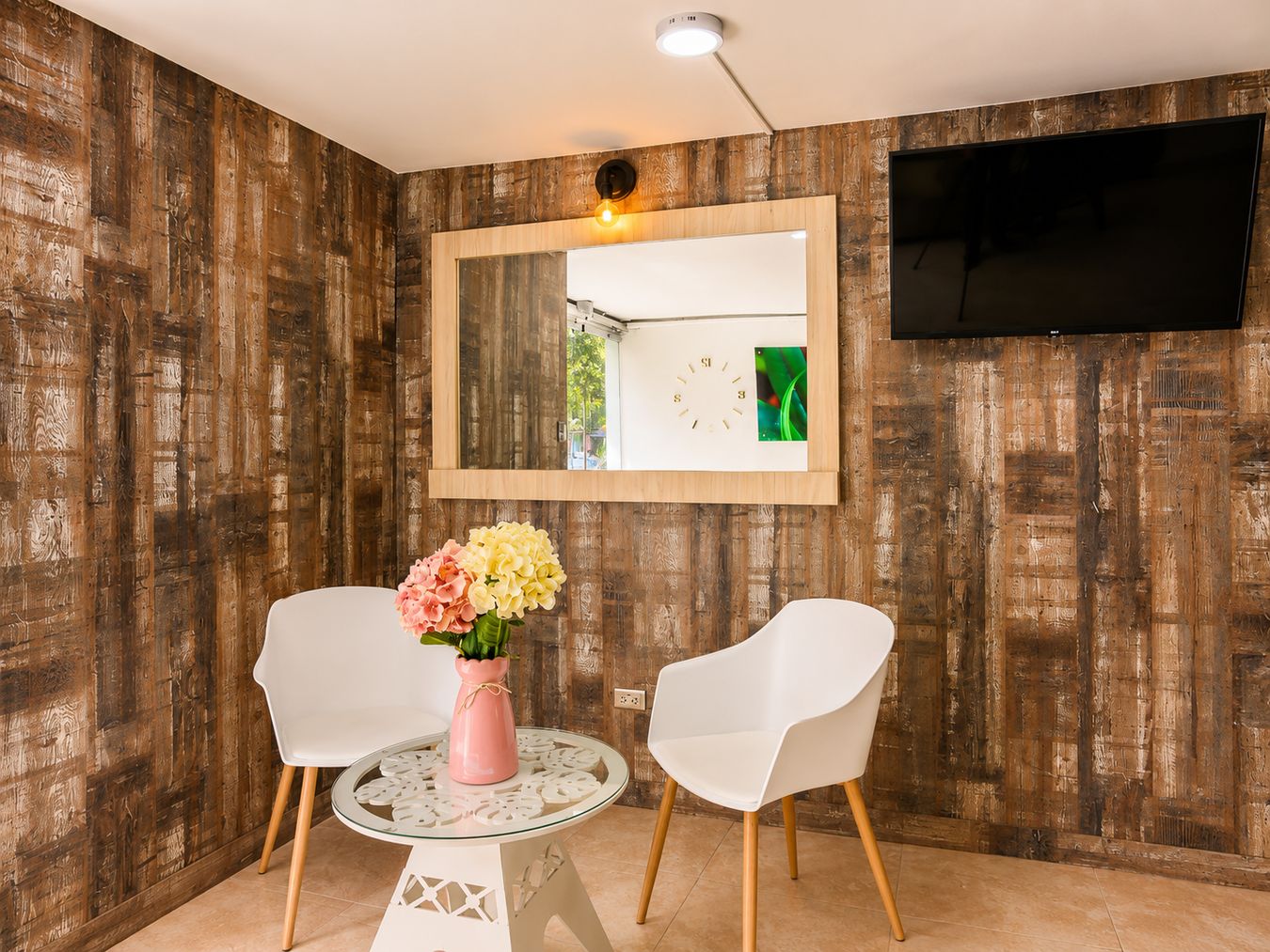 Quarto com paredes de madeira, mesa de vidro redonda com uma cadeira branca, um vaso rosa com flores e um espelho fixado acima da mesa.