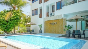 Piscina com borda azulejada, cercada por espreguiçadeiras e guarda-sóis, e um edifício com o letreiro 'Cocomo'.