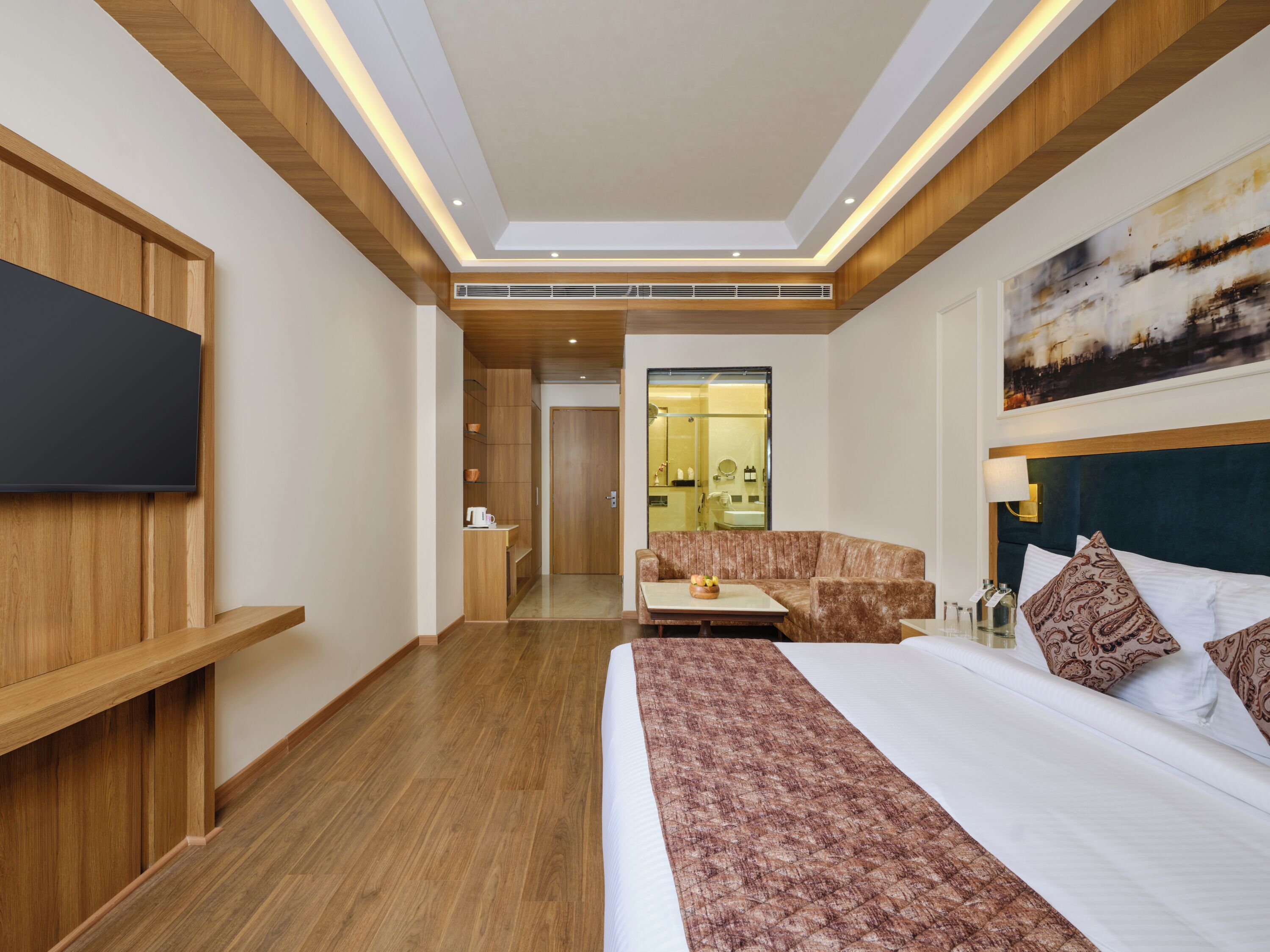 Quarto de hotel moderno com uma cama grande, um sofá, uma televisão de tela plana e um banheiro visível através de uma porta de vidro.