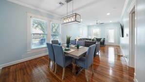 Uma área de refeições com uma mesa e cadeiras de madeira, um lustre pendurado e vista para a cozinha ao fundo.
