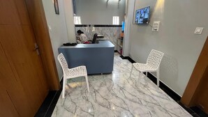 Uma área de recepção com piso de mármore, um balcão de atendimento, duas cadeiras brancas e uma televisão na parede.