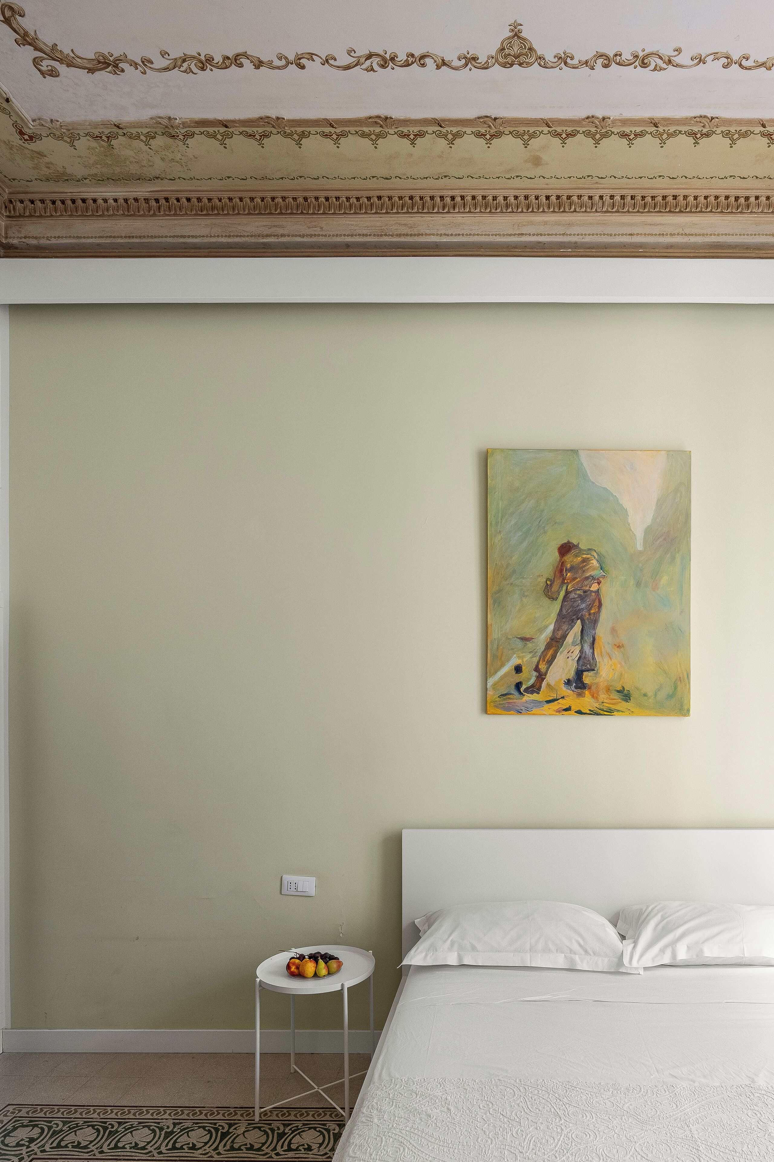 Um quarto com cama, uma pintura, uma mesinha com frutas e um tapete estampado.