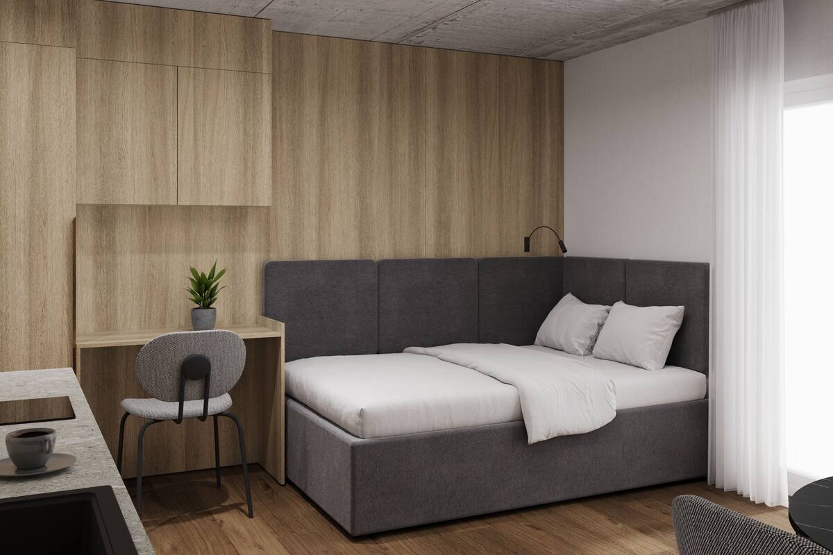 Quarto moderno com sofá-cama cinzento, uma pequena escrivaninha com cadeira, um vaso com planta e vista para o exterior através de uma janela com cortinas brancas.