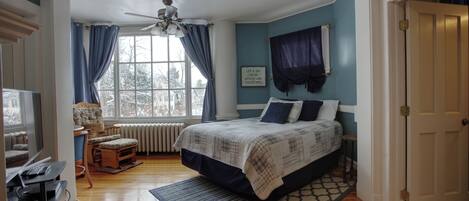Quarto com cama, escrivaninha, cadeira, janela com cortinas e ventilador de teto.