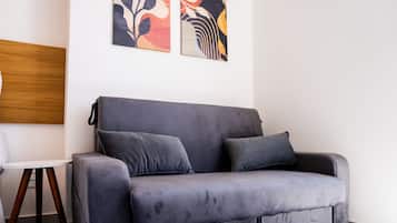 Um sofá cinzento com duas almofadas, uma mesa branca com base de madeira e arte abstrata na parede.