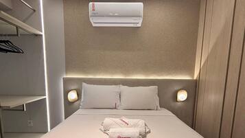 Quarto de hotel com cama, duas luminárias laterais, unidade de ar condicionado e toalha dobrada sobre a cama.