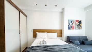 Quarto de hotel com cama, guarda-roupa, mesa de cabeceira, comando remoto, sofá cinzento e duas obras de arte penduradas na parede.