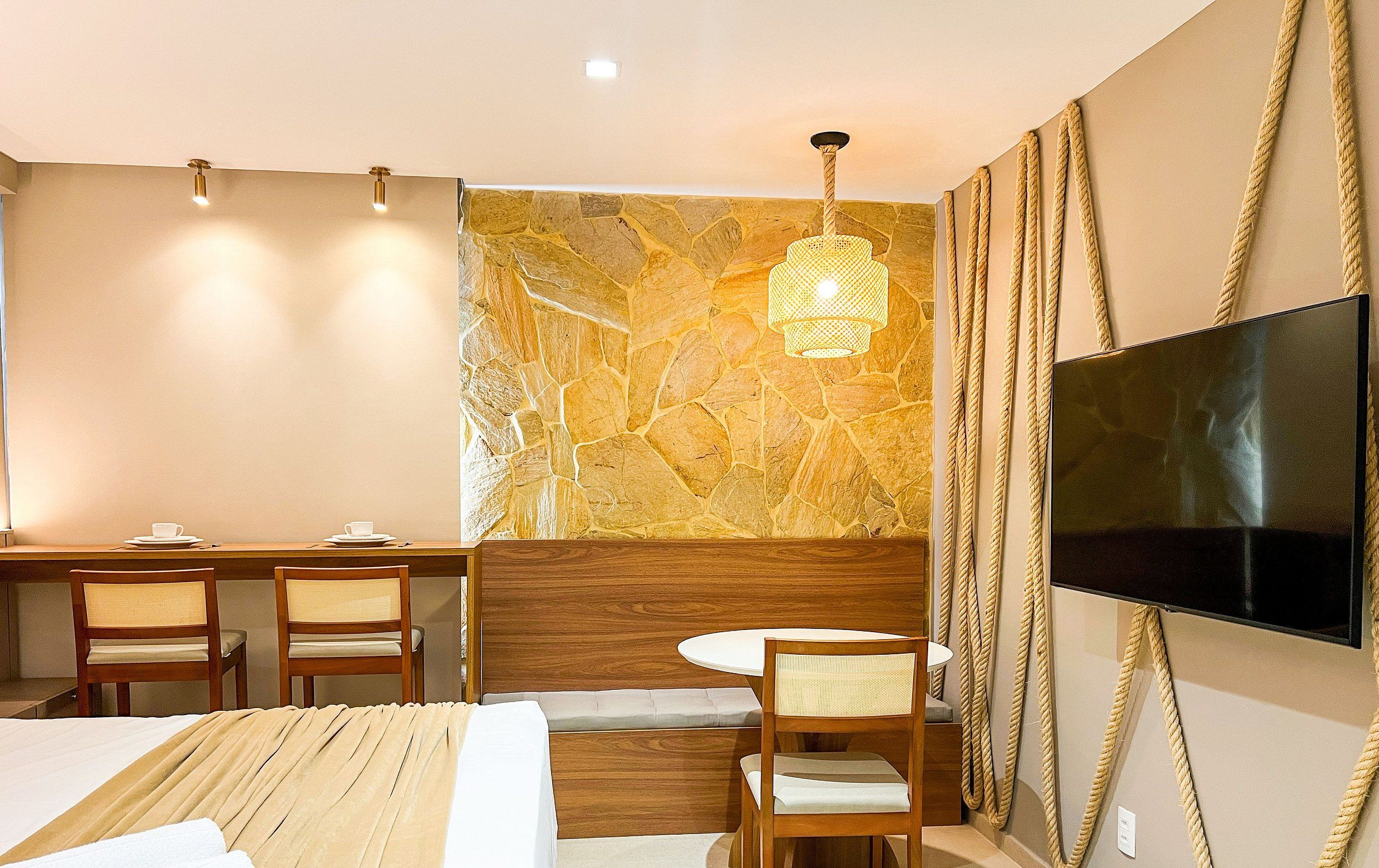 Quarto de hotel com cama, escrivaninha com cadeiras, televisão e parede de pedra.