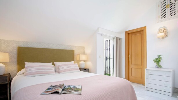 Quarto de hotel bem arrumado com uma cama grande, abajures, uma pequena mesa de cabeceira, cômoda branca e uma porta que dá para uma varanda.