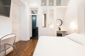 Quarto com cama, poltrona, escrivaninha, espelho e banheiro visível por uma porta aberta.