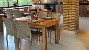 Uma ampla sala de jantar com mesas e cadeiras de madeira, um pilar de tijolos e uma cozinha ao fundo.