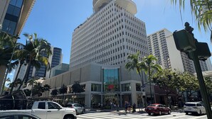 Um edifício moderno com um topo circular característico, rodeado por palmeiras e outras construções urbanas.