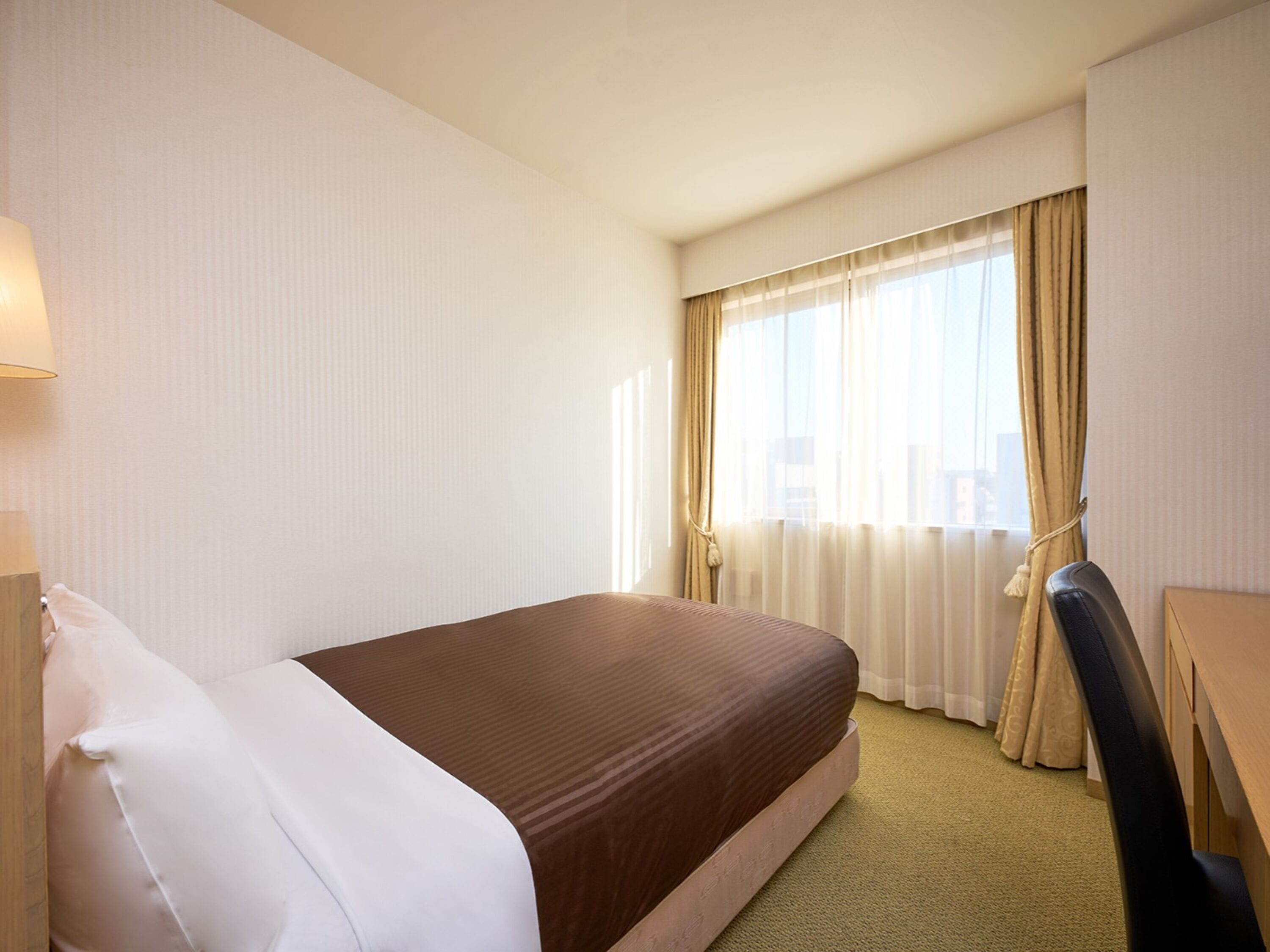 Quarto de hotel com cama, escrivaninha, cadeira e janela com cortinas.