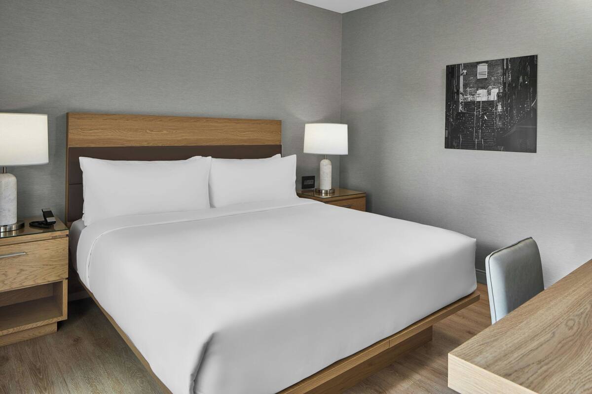 Quarto de hotel moderno com uma cama grande, cabeceira de madeira, duas mesas de cabeceira com luminárias, uma escrivaninha e uma cadeira.