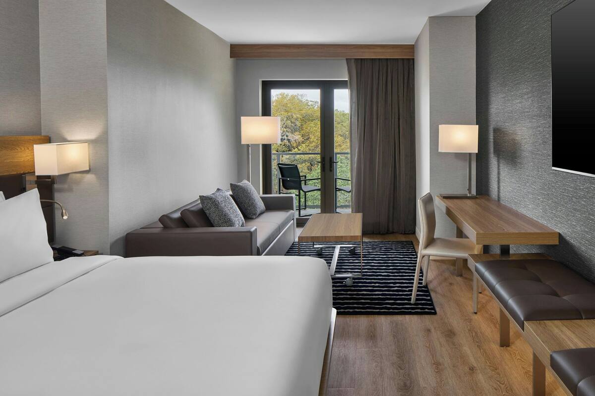 Quarto de hotel moderno com uma cama grande, um sofá, uma escrivaninha e uma varanda com vista para árvores.