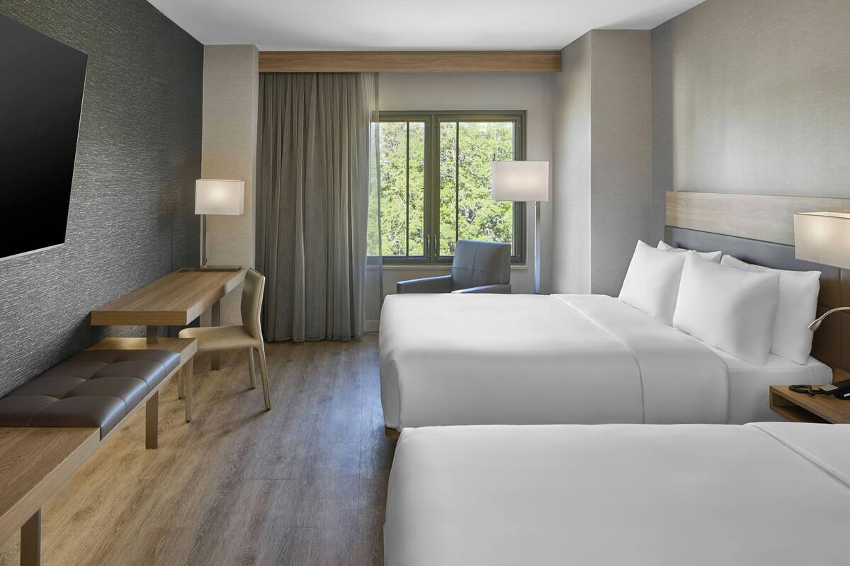 Quarto de hotel com uma cama grande, uma escrivaninha com cadeira, um banco, duas luminárias de cabeceira e uma televisão na parede.