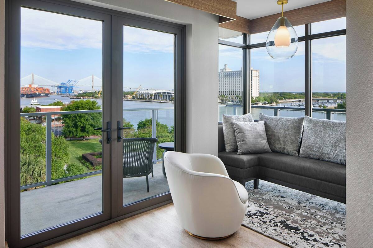 Uma sala de estar moderna com sofá e poltrona, grandes janelas com vista para uma ponte e para a água, e um lustre.