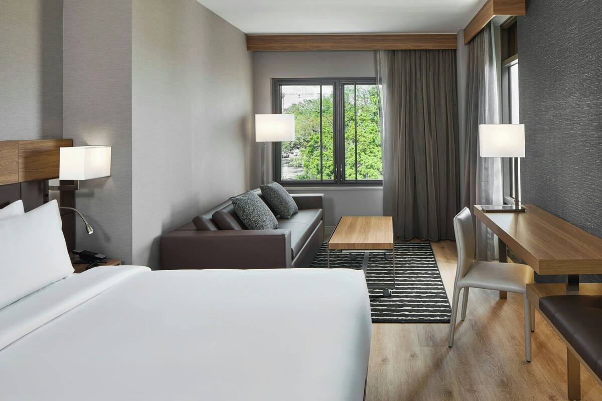 Quarto de hotel com cama, sofá, escrivaninha e janela com cortinas.