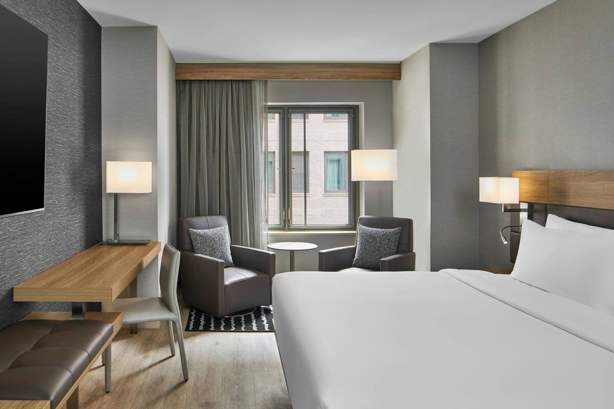 Quarto de hotel moderno com uma cama grande, duas poltronas, uma mesa pequena e uma escrivaninha.