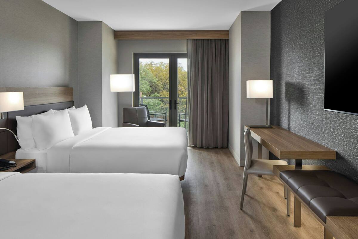 Quarto de hotel com duas camas, uma escrivaninha, uma cadeira, televisão e varanda com cortinas.