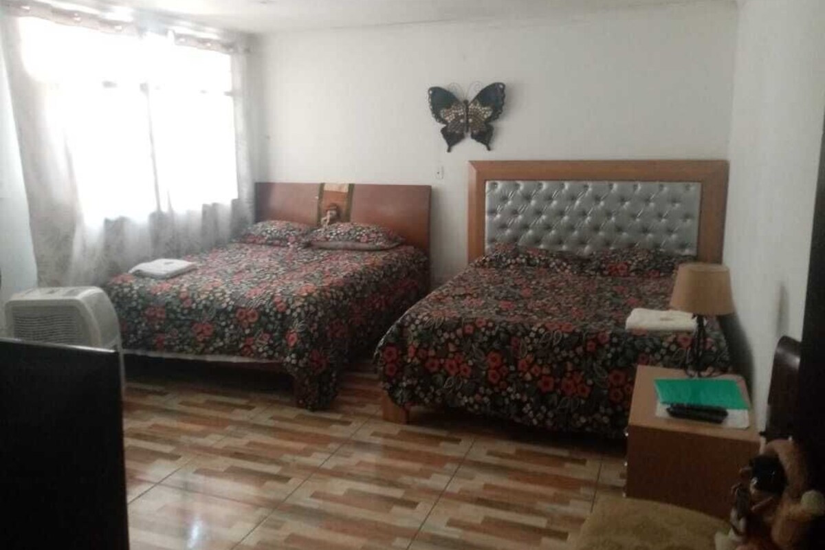 Quarto com duas camas, um ornamento de parede em formato de borboleta, abajur e ventilador.