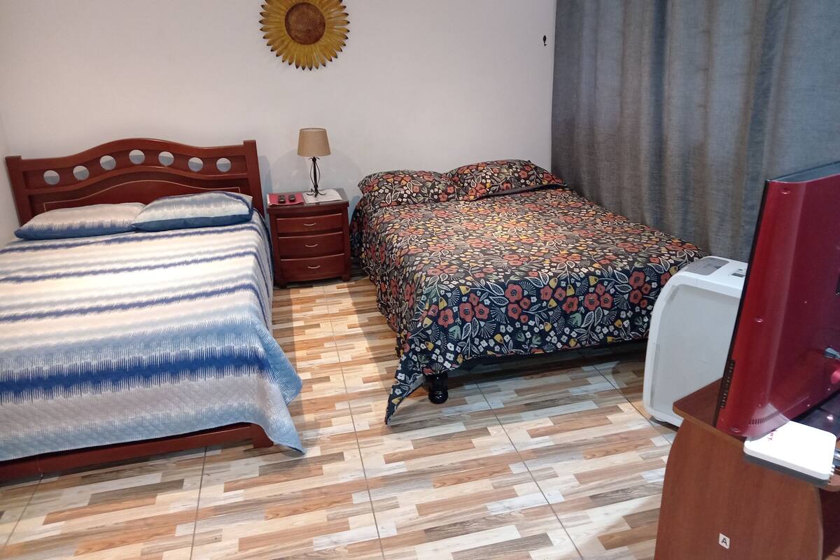 Quarto com duas camas, mesa de cabeceira, luminária, televisão e decoração de sol na parede.