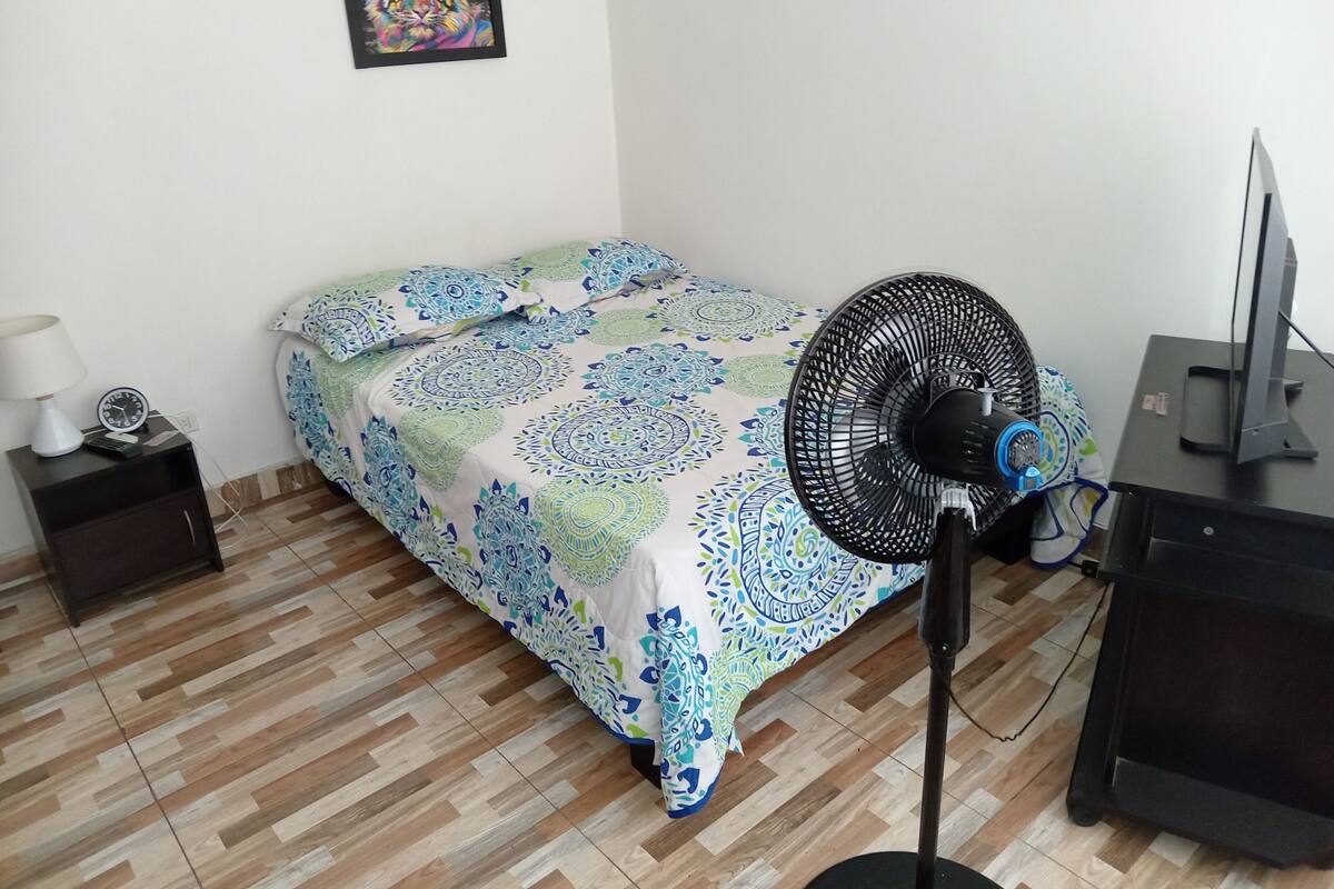 Quarto com cama, um ventilador, mesa de cabeceira com luminária, relógio e televisão sobre um suporte.