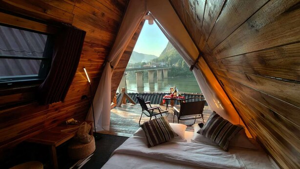 Uma cabana acolhedora com uma cama, uma mesa e vista para um rio e uma ponte.