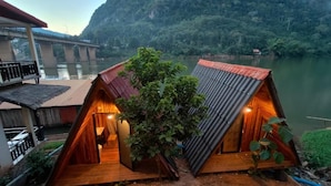 Uma cabana de madeira com telhado inclinado, um pequeno alpendre e vista para uma ponte e colinas arborizadas.