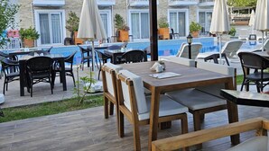 Uma área externa com mesas e cadeiras de madeira, piscina e guarda-sóis.