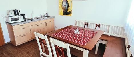 Uma área de refeições com mesa de madeira, cadeiras brancas e um banco. Há uma cozinha com micro-ondas e chaleira. Um quadro com um vaso e flores está pendurado na parede.