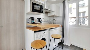 Uma cozinha compacta com micro-ondas, máquina de café e máquina de lavar roupa. Há dois bancos e uma janela com vista para o exterior.