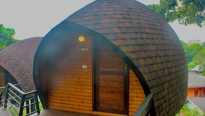Uma cabana de madeira em formato de cúpula, com porta de madeira e uma pequena escadaria que leva a um deck.