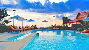 Piscina com espreguiçadeiras e guarda-sóis, cercada por palmeiras e um céu ao pôr do sol.