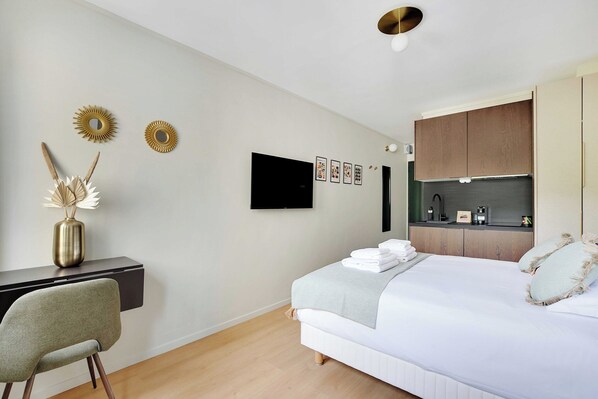 Quarto moderno com cama, escrivaninha e pequeno espaço de cozinha integrado.