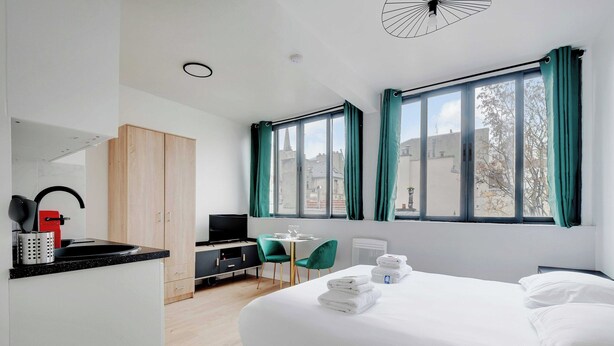 Quarto de hotel moderno com uma cama grande, uma escrivaninha com computador, uma mesa pequena com cadeiras e vista para a cidade através de grandes janelas.