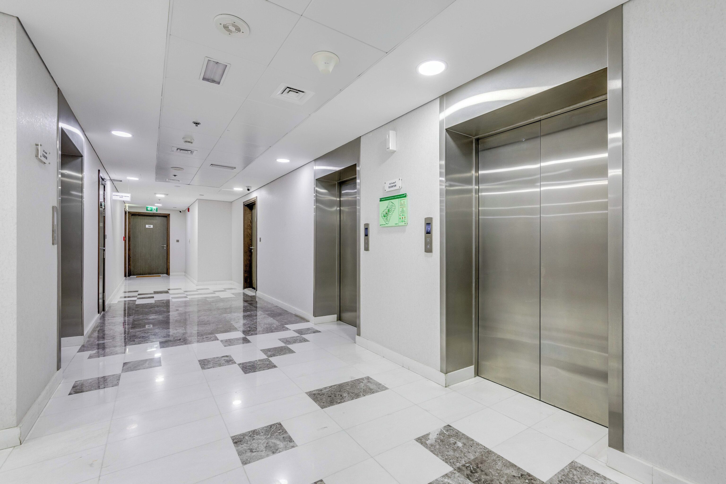 Um corredor limpo e moderno com piso xadrez, dois elevadores e uma porta no final.