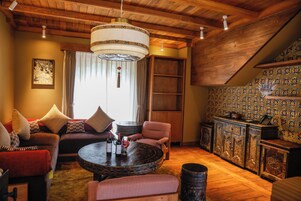Uma sala de estar aconchegante com teto de madeira, mesa de centro redonda com garrafas de vinho, sofá com almofadas e parede estampada.