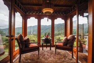 Quarto com teto de madeira, um candelabro, duas cadeiras com almofadas, uma mesa com garrafa e copo, e vista para uma paisagem montanhosa através de grandes janelas.