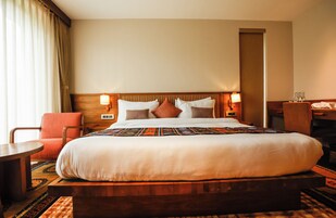 Quarto de hotel com uma cama grande, cabeceira de madeira, duas luminárias laterais, uma poltrona vermelha, uma mesinha e piso coberto por carpete.
