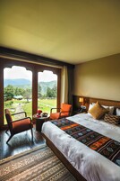 Quarto de hotel com cama grande, uma cadeira, uma mesinha com vaso e frutas, e vista para uma paisagem montanhosa pelas janelas.