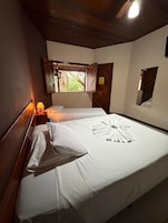 Quarto de hotel com duas camas, mesa de cabeceira com luminária, janela com cortinas e teto de madeira.