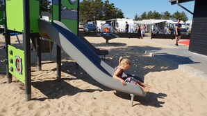 Um parquinho com escorrega, areia e uma área coberta ao fundo.