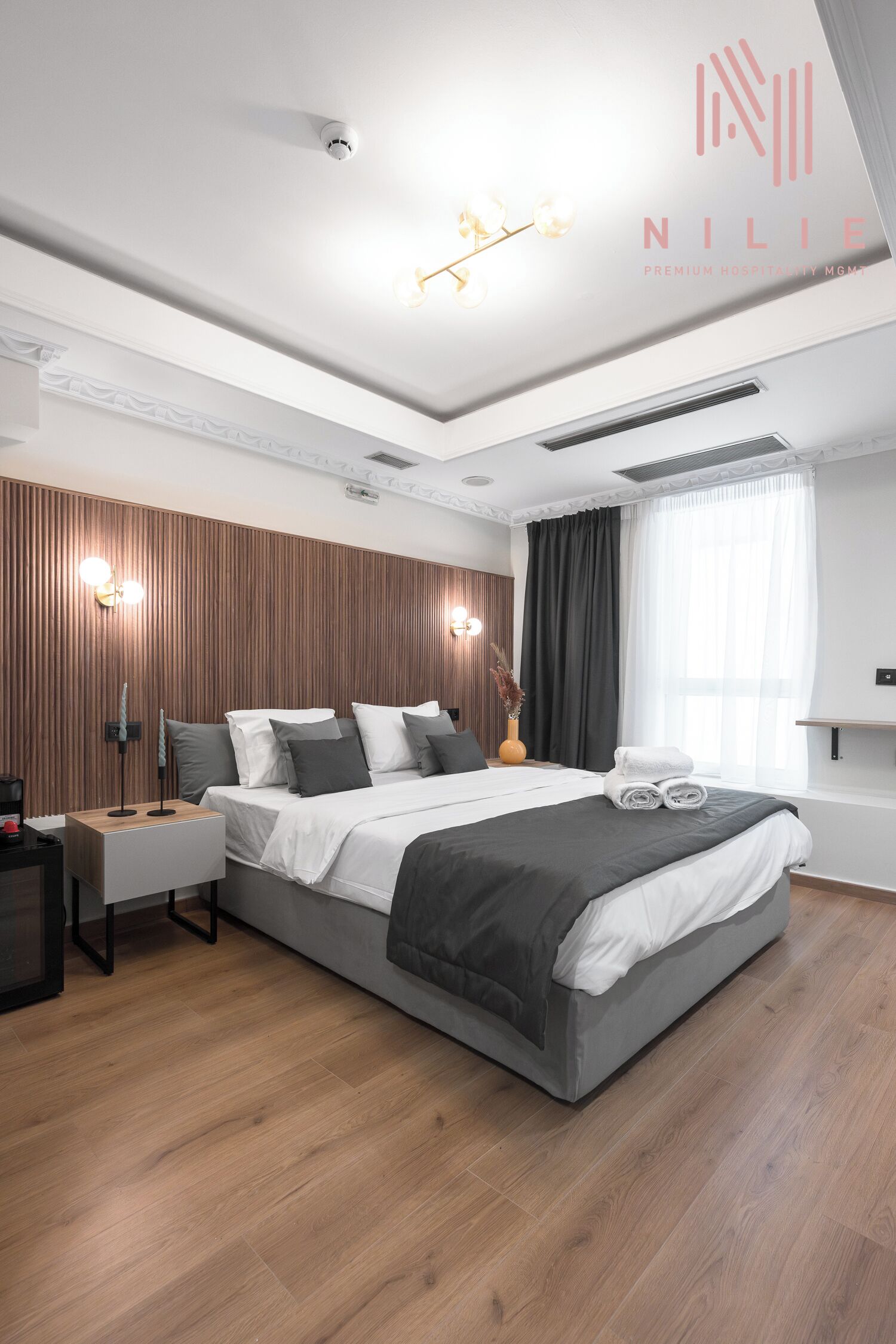 Quarto de hotel moderno com uma cama grande, mesas de cabeceira, escrivaninha e uma janela ampla com cortinas.