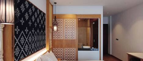Quarto de hotel moderno com uma cama grande, luminária de cabeceira, parede com padrão, pequena escrivaninha e banheiro visível por uma porta aberta.